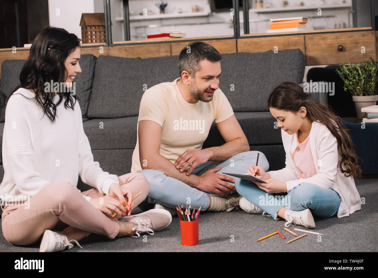 happy parents sitting on floor near cute daughter drawing on paper ...