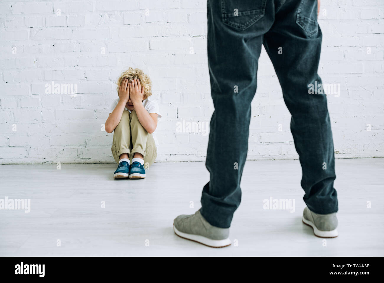 cropped view of father holding belt and scared son sitting on floor ...