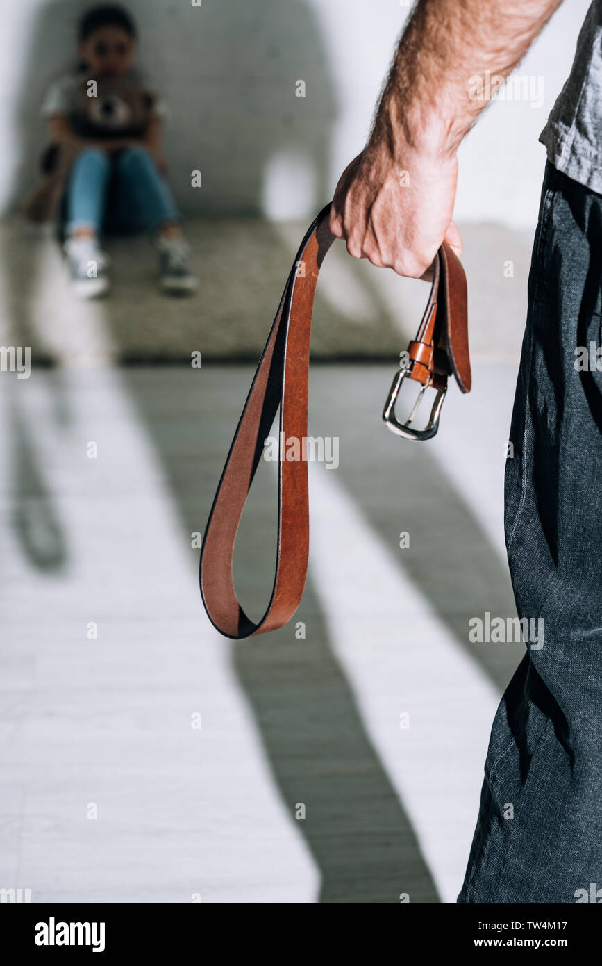 cropped view of father holding belt and scared daughter sitting on ...