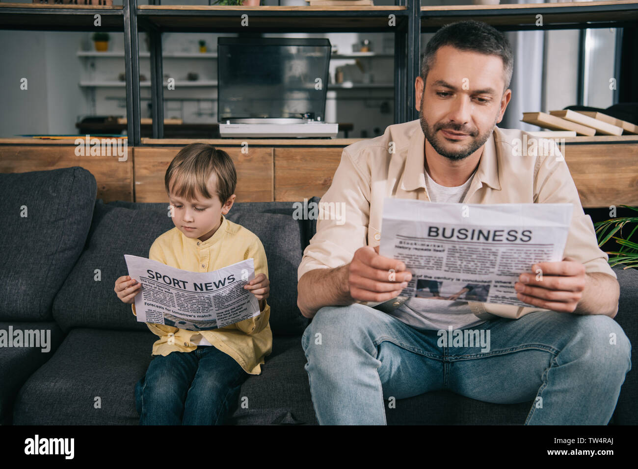 concentrated father and son reading newspapers while sitting on sofa at ...
