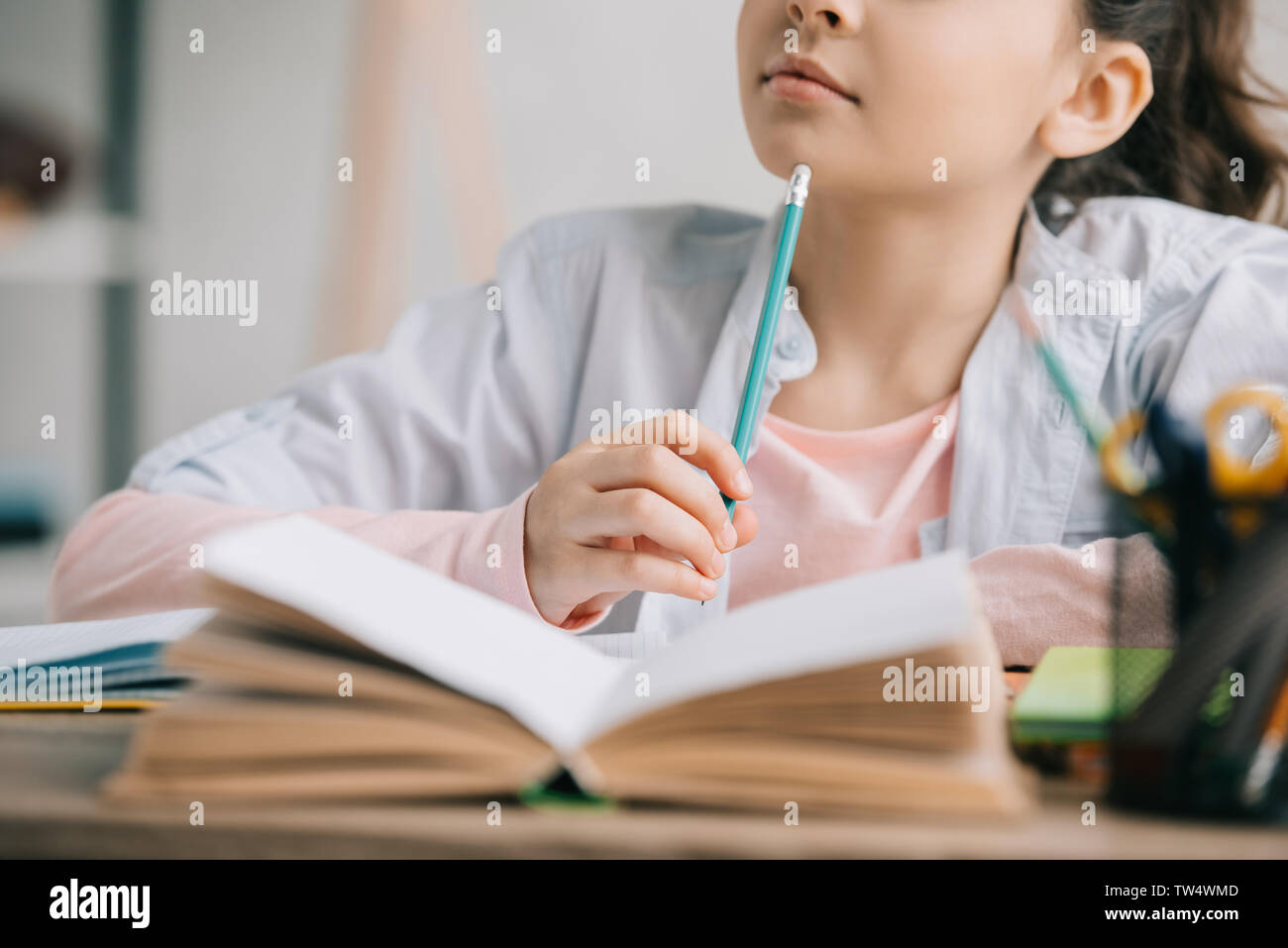 partial view of schoolkid holding pencil while sitting at desk near ...