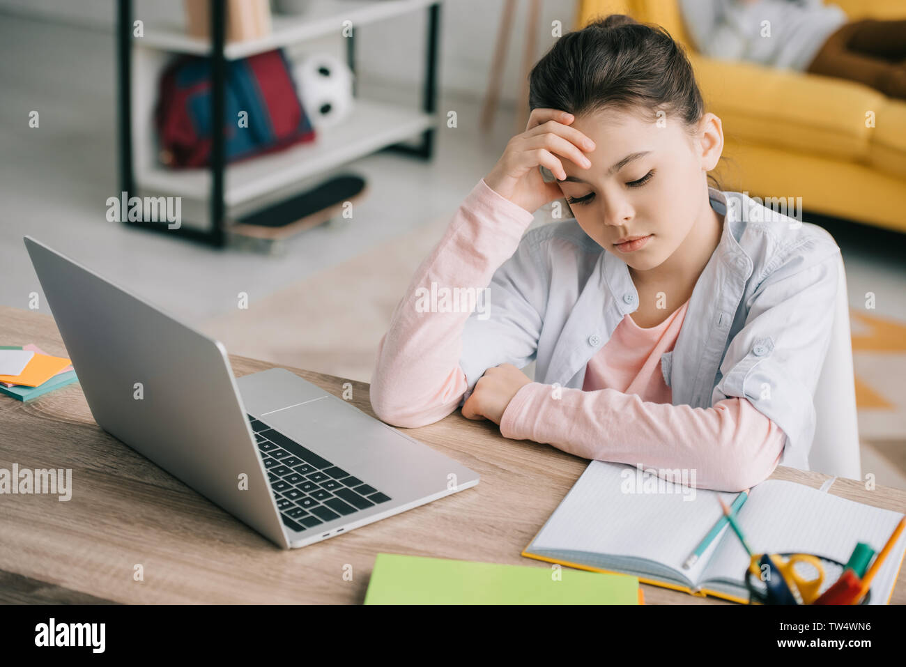 exhausted schoolchild sitting at desk near laptop and copy book while doing schoolwork at home ...