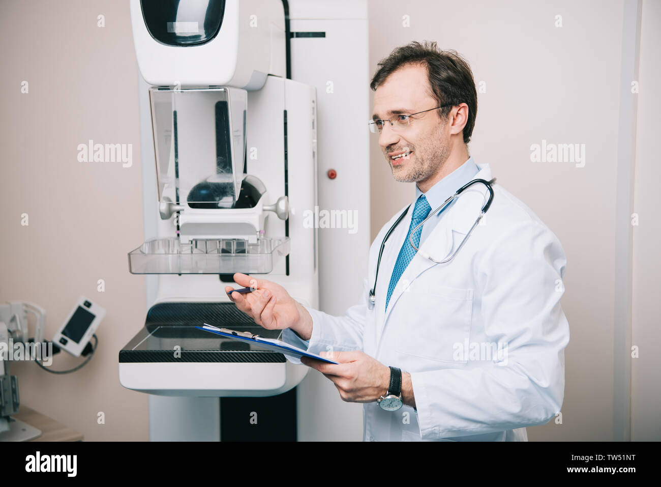 smiling radiographer standing near x-ray machine and holding clipboard ...