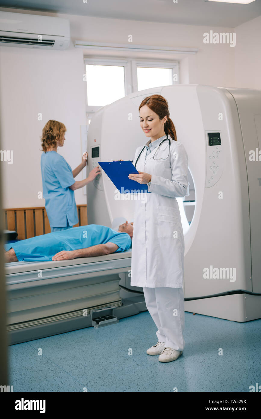 smiling radiographer writing on clipboard while assistant standing near ...