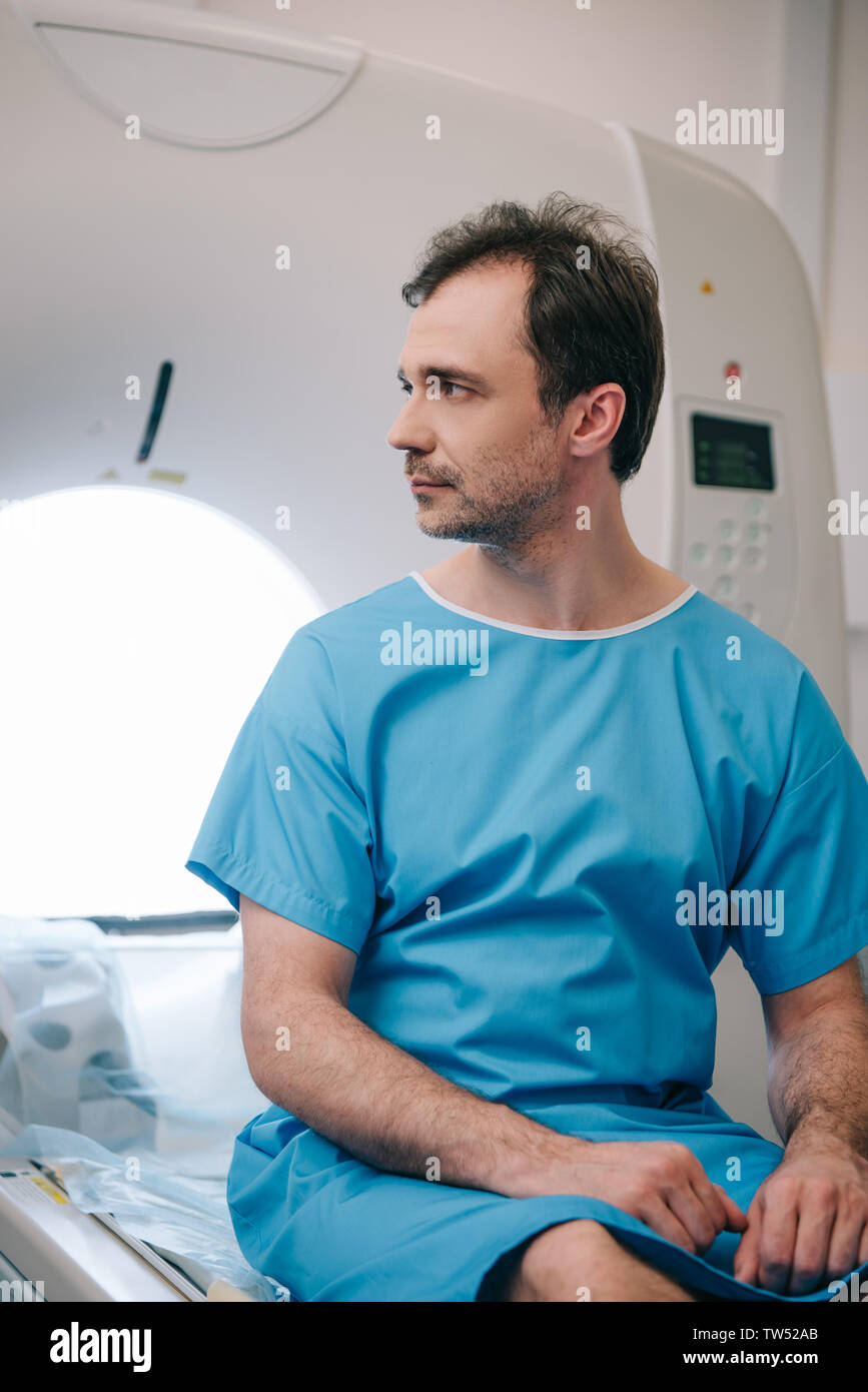 handsome man sitting on ct scanner bed in hospital and looking away ...