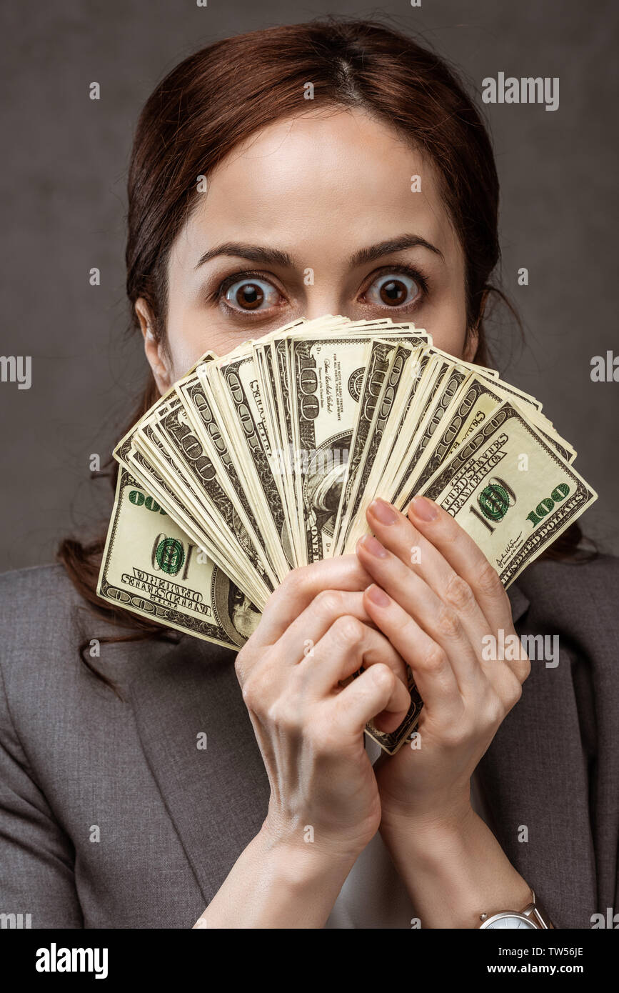 shocked brunette woman covering face with dollar banknotes on grey ...