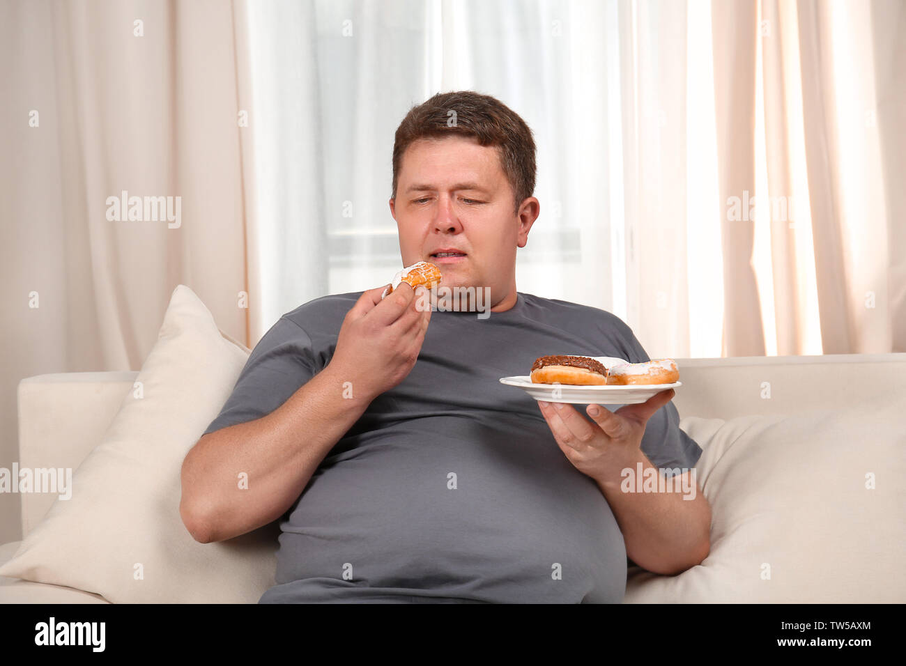 Overweight young man eating sweets on sofa at home Stock Photo - Alamy