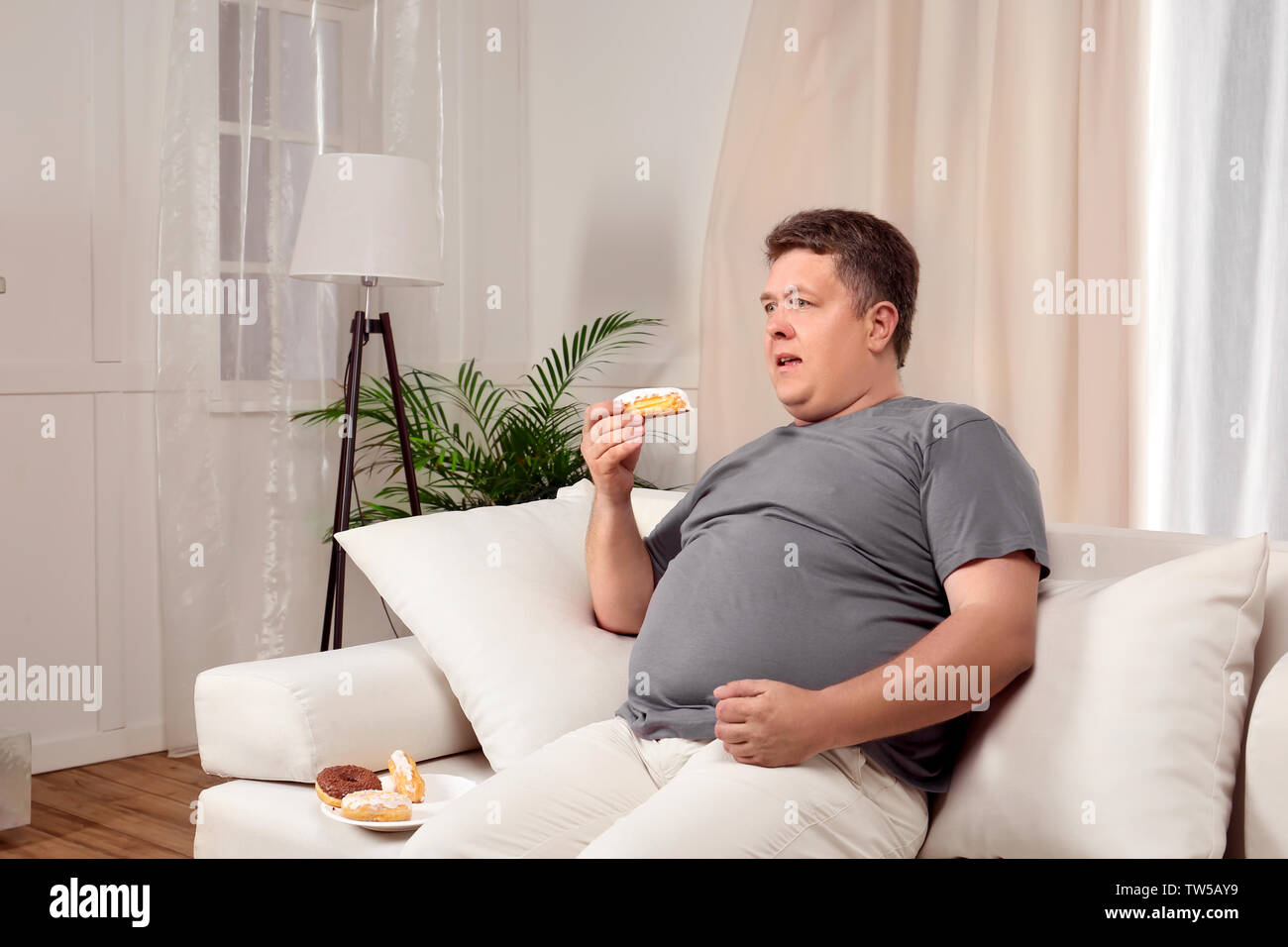 Overweight young man eating sweets on sofa at home Stock Photo - Alamy
