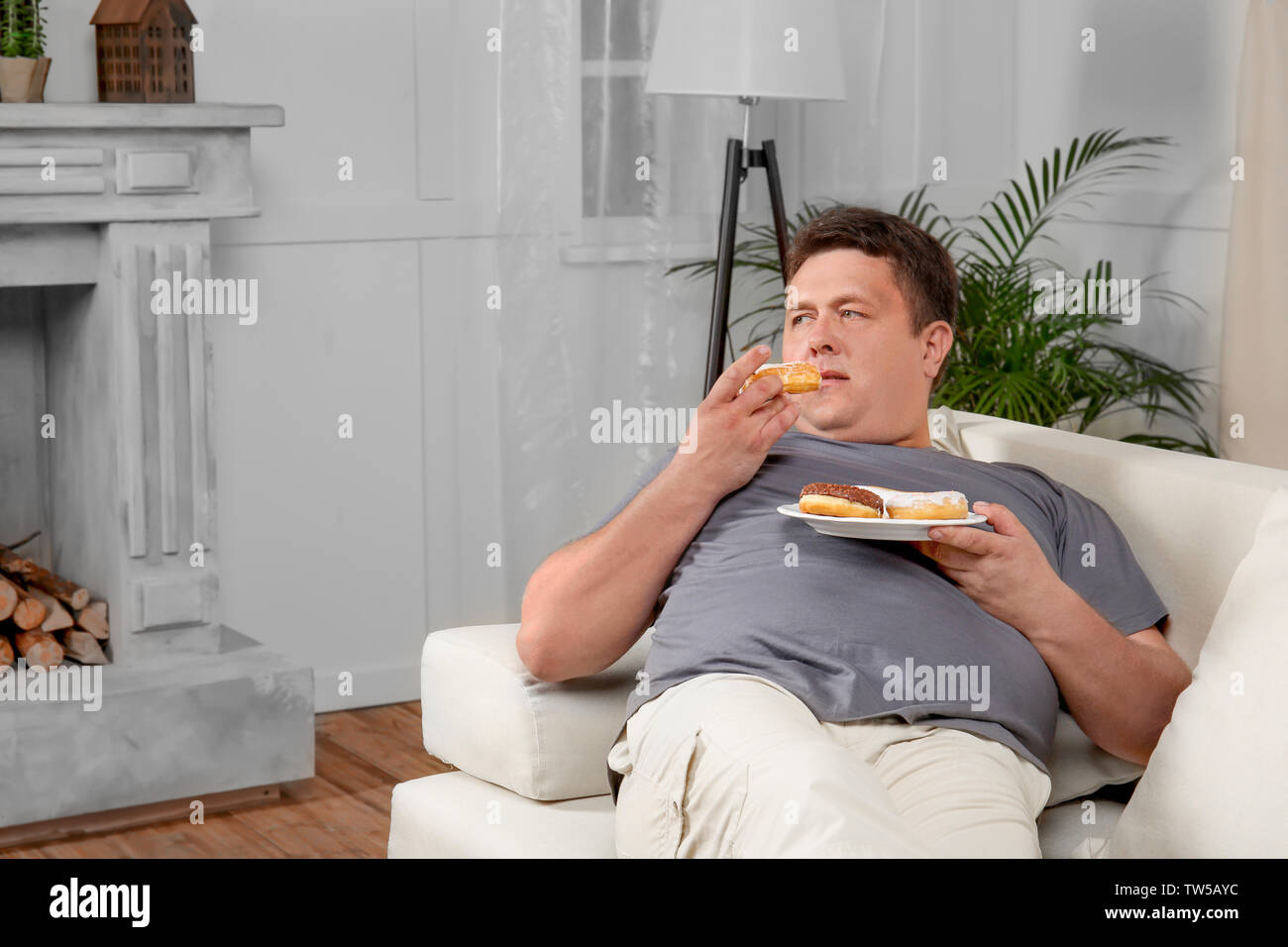 Overweight young man eating sweets on sofa at home Stock Photo - Alamy