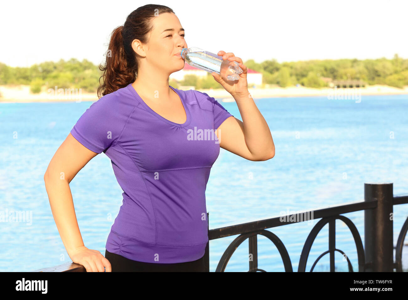 Overweight young woman in sportswear drinking water near river. Weight ...
