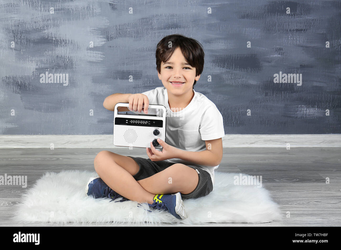 Little boy listening to radio while sitting on floor against grey wall ...