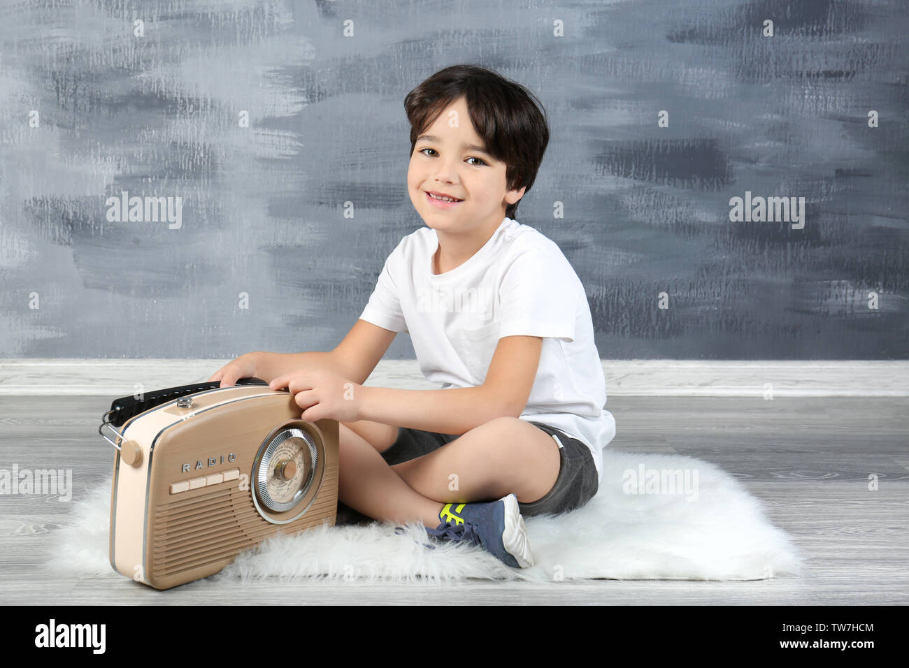 Little boy listening to radio while sitting on floor against grey wall ...