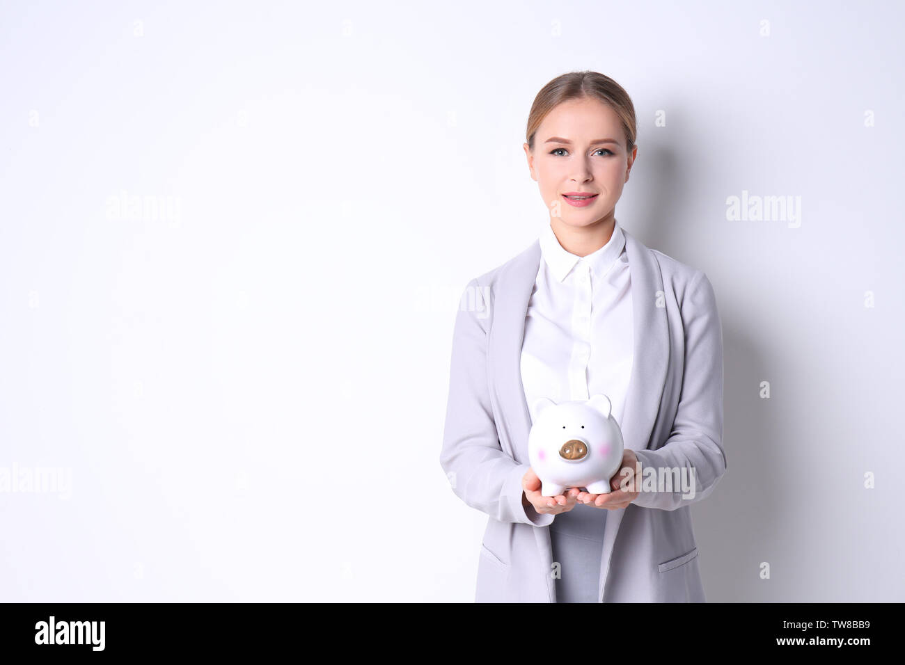 Young woman in formal clothes holding piggy bank on white background ...