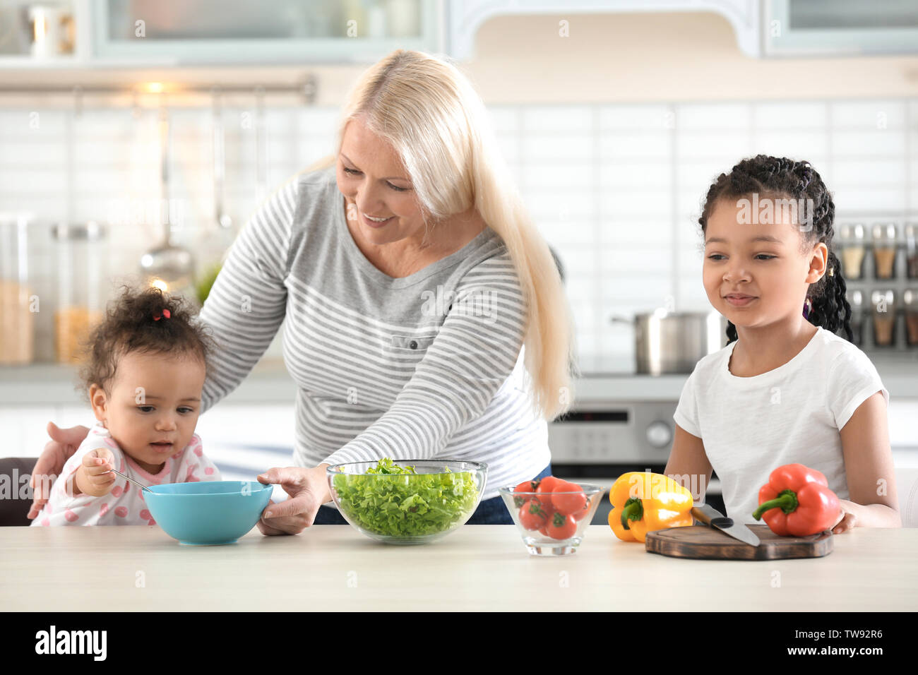 Female mature nanny feeding little African-American girl in kitchen ...