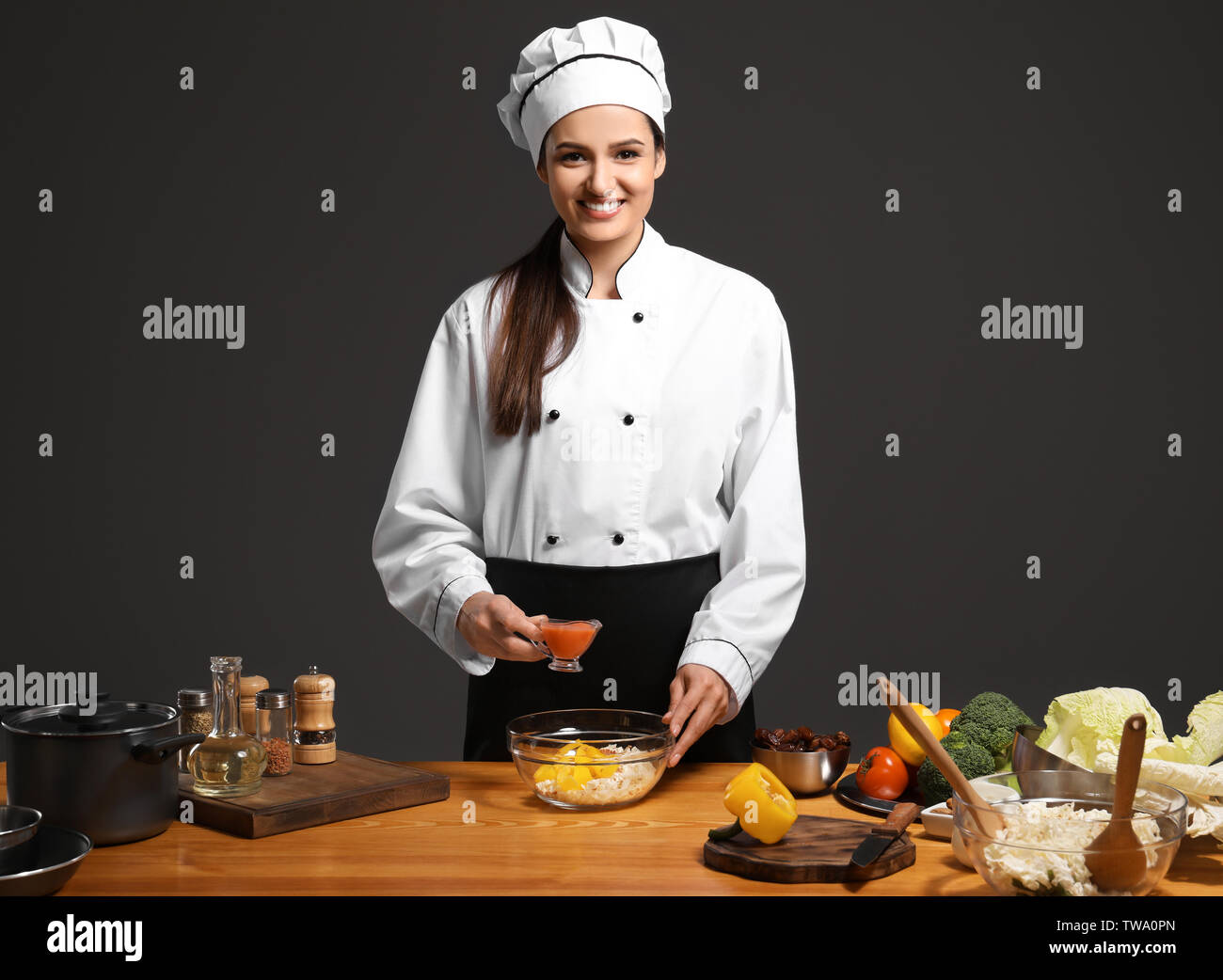 Beautiful female chef adding sauce to salad at table against dark ...