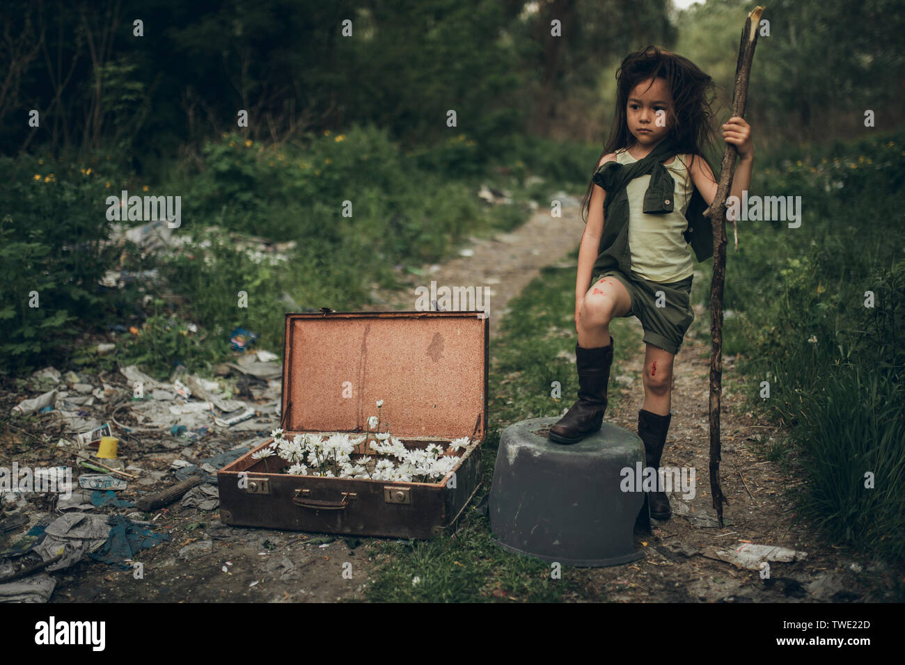 A homeless girl is standing on a garbage dump next to a suitcase with ...