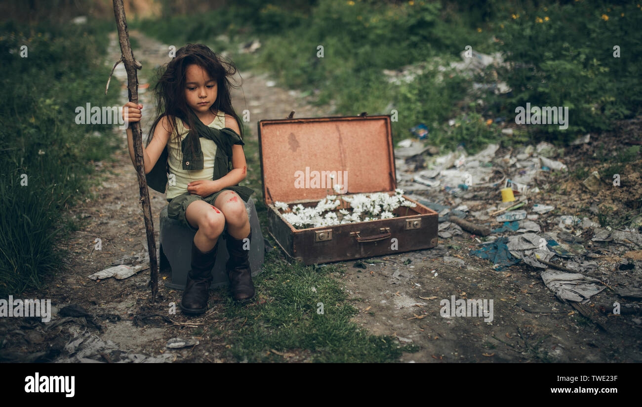 A homeless girl is sitting on a garbage dump next to a suitcase with ...