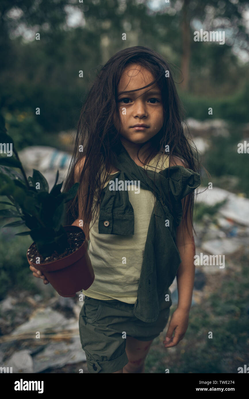 A homeless girl is standing on a garbage dump with a houseplant in a ...