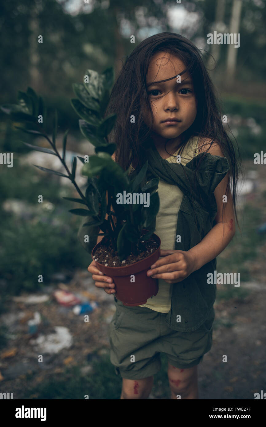 A homeless girl is standing on a garbage dump with a houseplant in a ...
