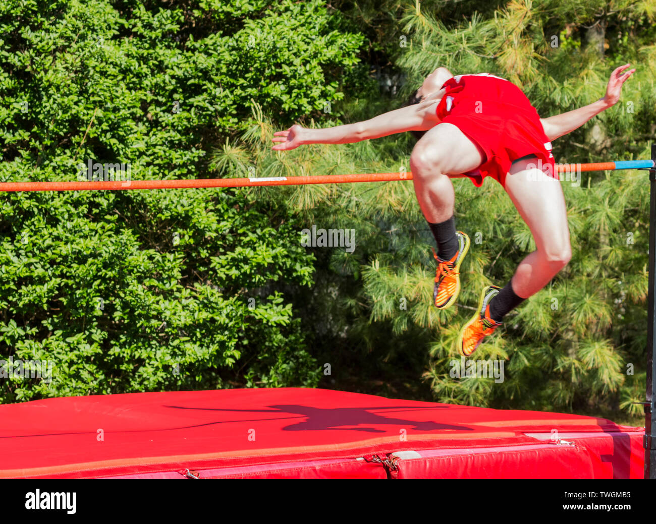 A male high school high jumper is over the bar with trees in the ...
