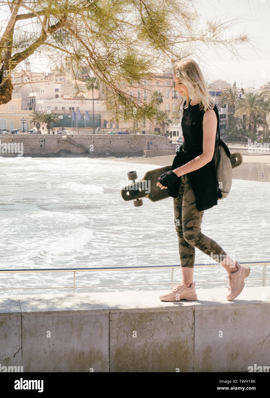Pretty young girl with a skateboard in the hands walking along seafront ...