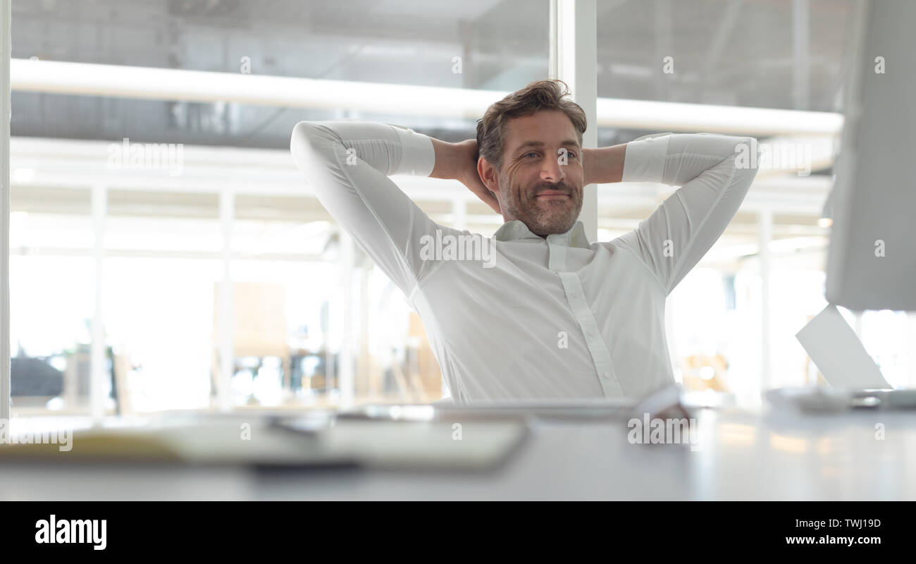 Business male executive with hands behind hand sitting at desk in a ...