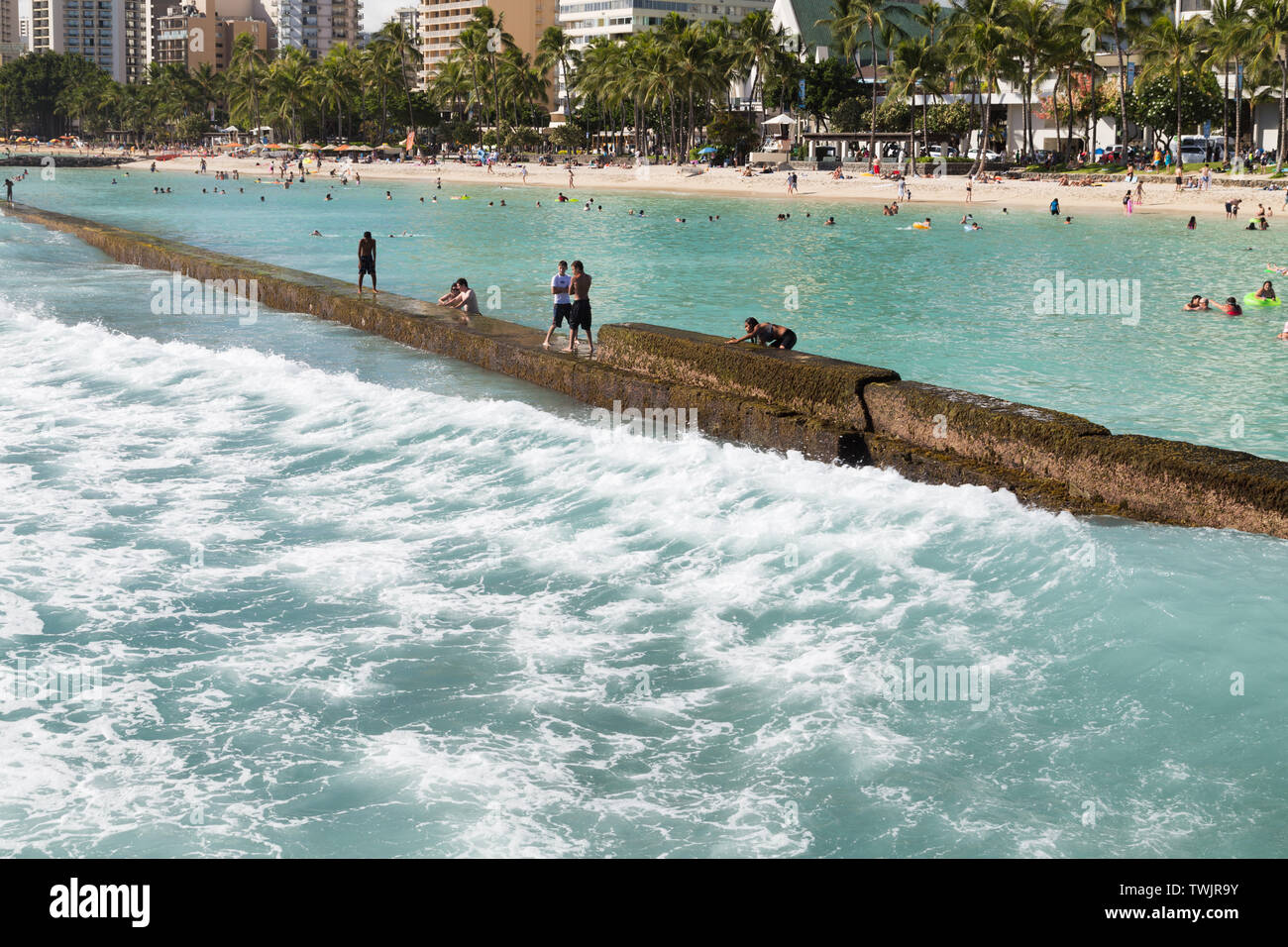 The incoming waves hit the seawall while tourists and locals walk on it ...