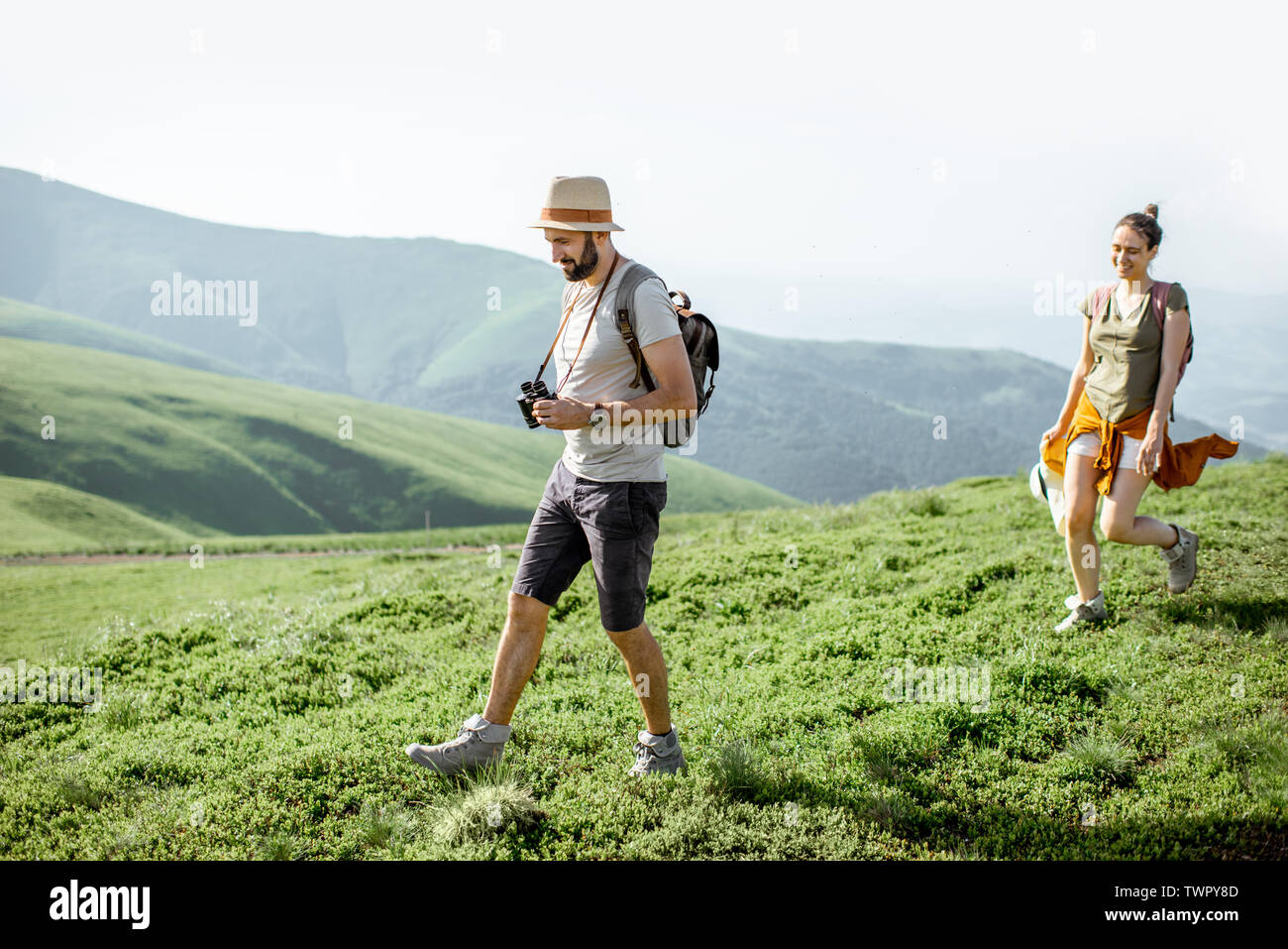 Beautiful couple walking with backpacks on the green meadow, having fun while traveling in the ...