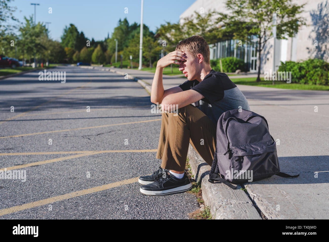 Upset teenager resting his head on his hand as he sits on the curb in ...