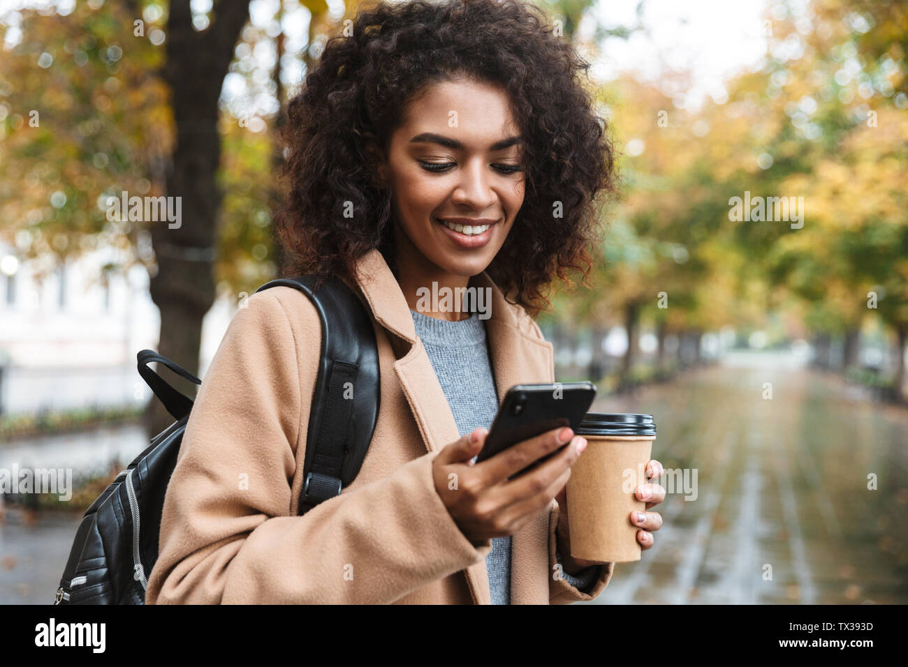 Beautiful young african woman wearing coat walking outdoors at the park, carrying backpack ...