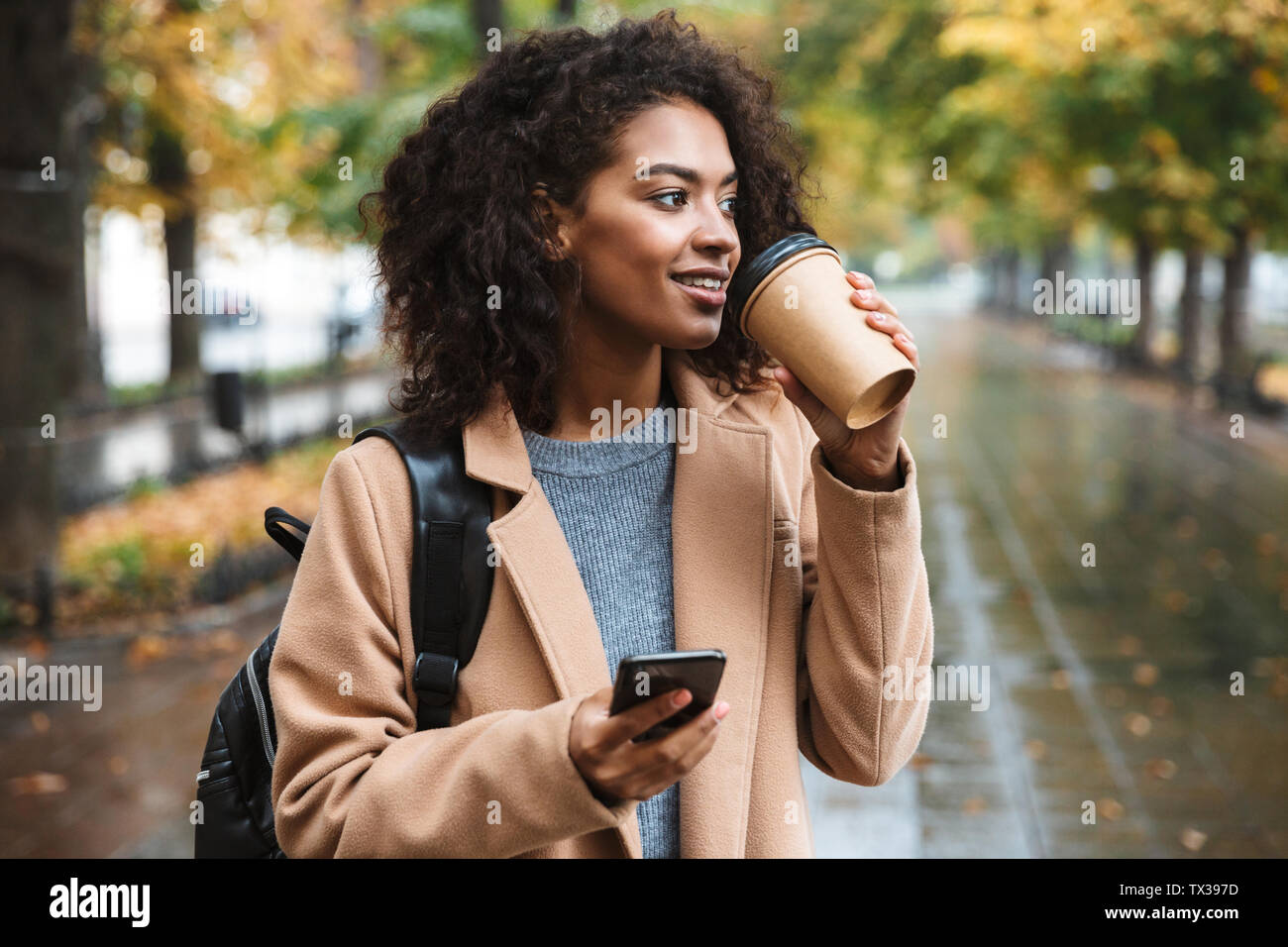 Beautiful young african woman wearing coat walking outdoors at the park, carrying backpack ...