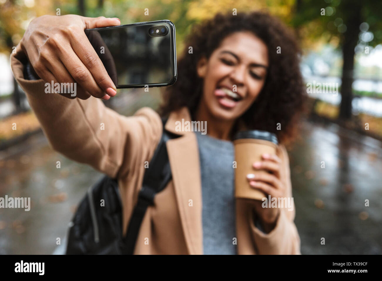 Beautiful young african woman wearing coat walking outdoors at the park, carrying backpack ...