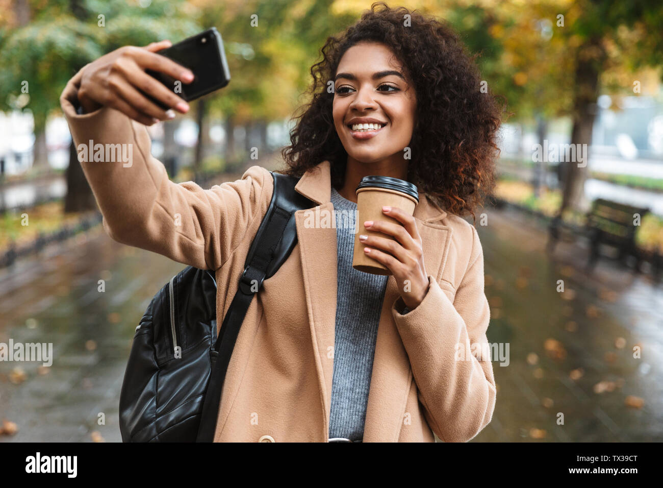 Beautiful young african woman wearing coat walking outdoors at the park, carrying backpack ...