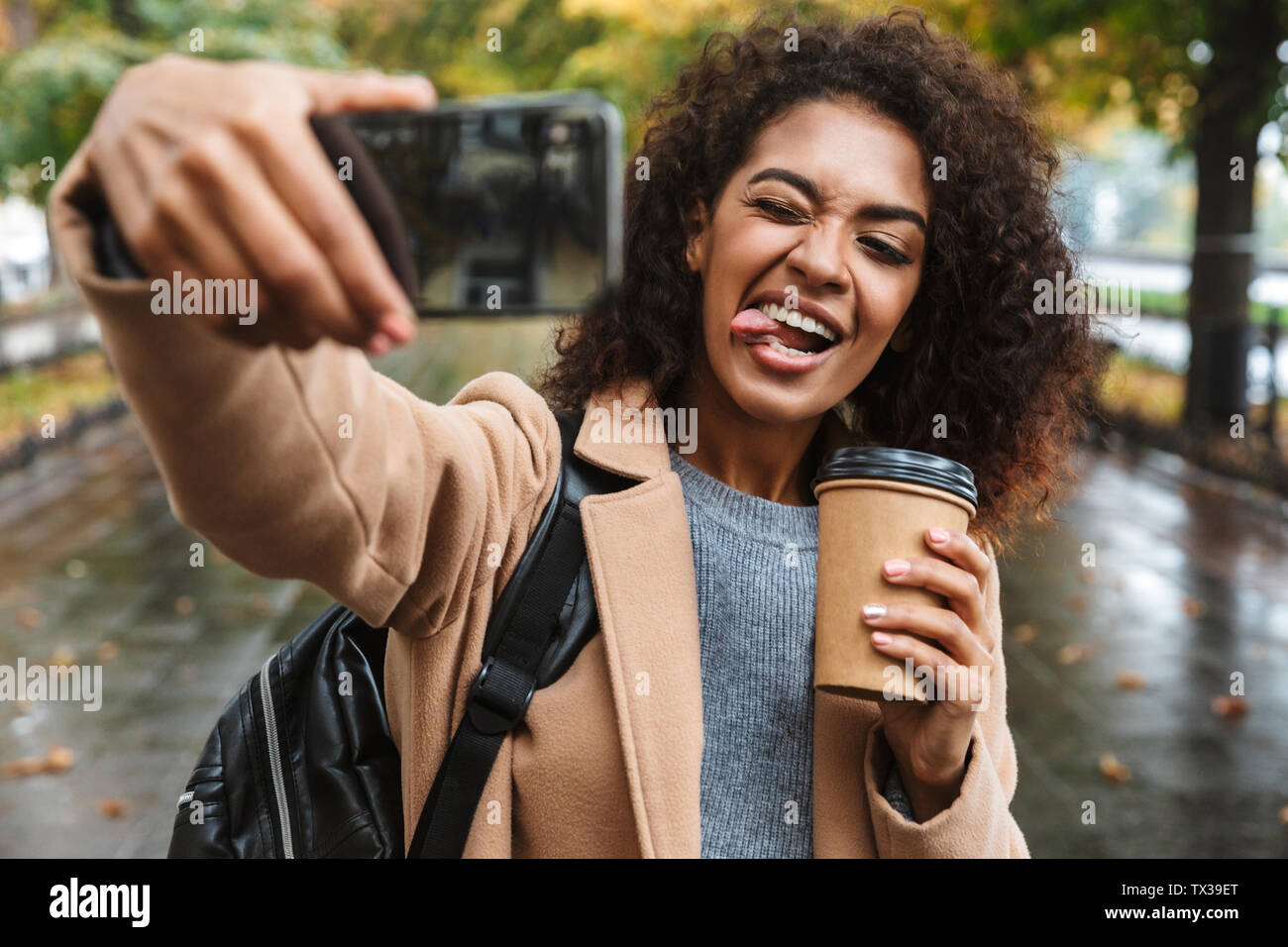 Beautiful young african woman wearing coat walking outdoors at the park, carrying backpack ...