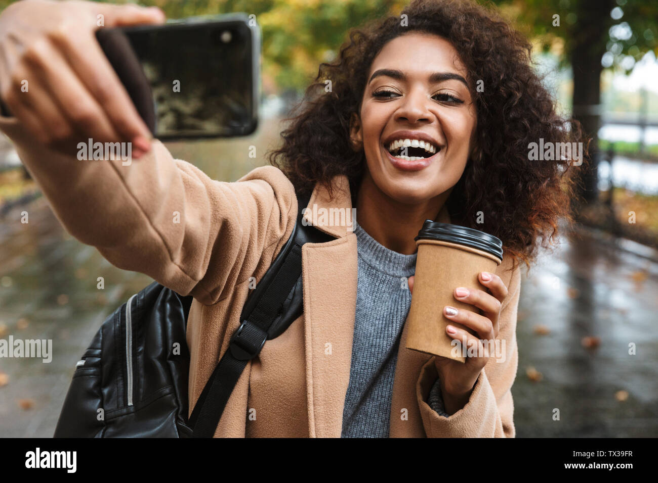 Beautiful young african woman wearing coat walking outdoors at the park, carrying backpack ...
