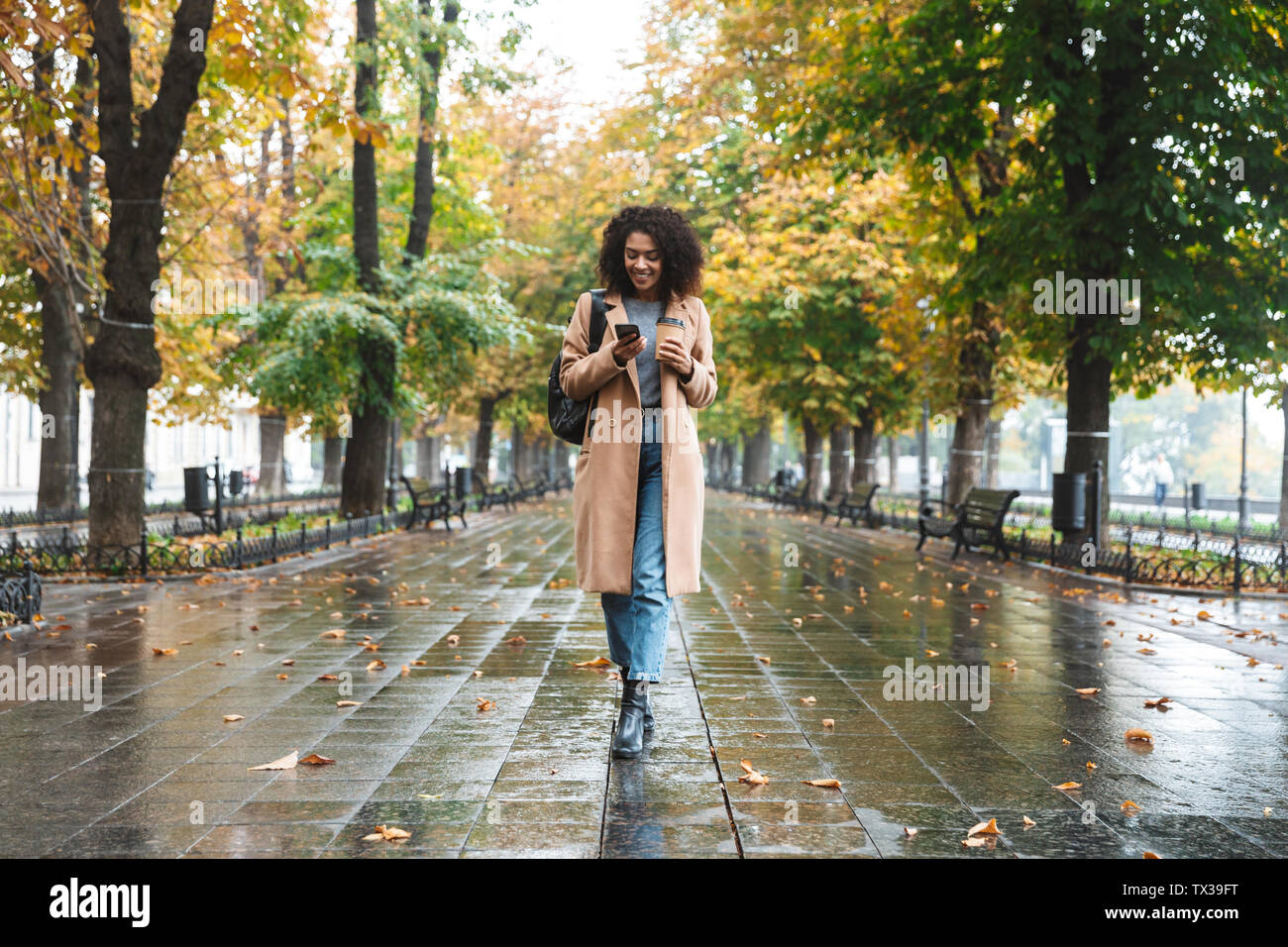 Beautiful young african woman wearing coat walking outdoors at the park, carrying backpack ...