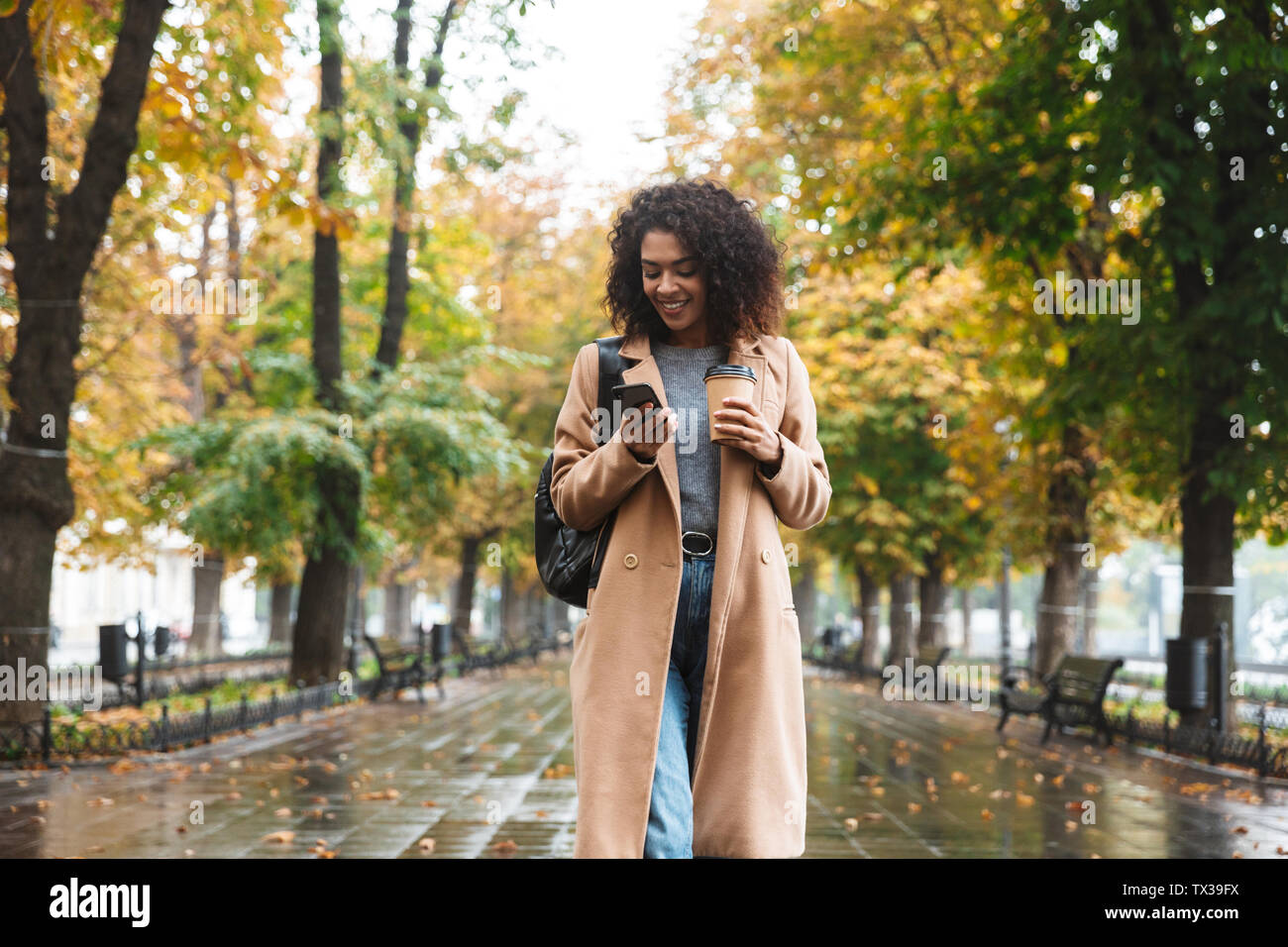 Beautiful young african woman wearing coat walking outdoors at the park, carrying backpack ...