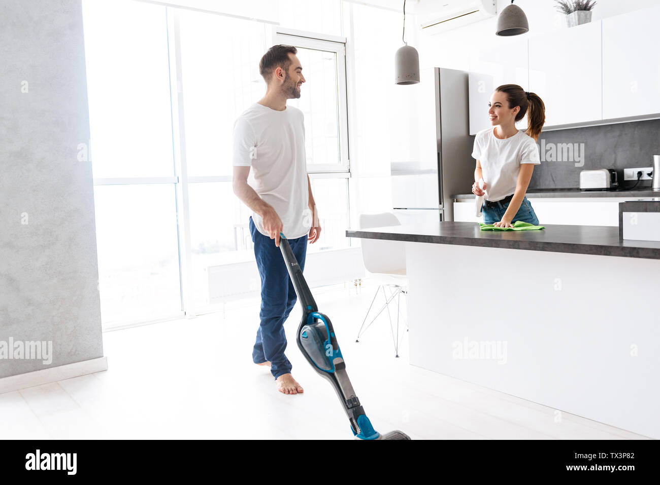 Happy young couple doing general clean at the kitchen, man vacuuming ...