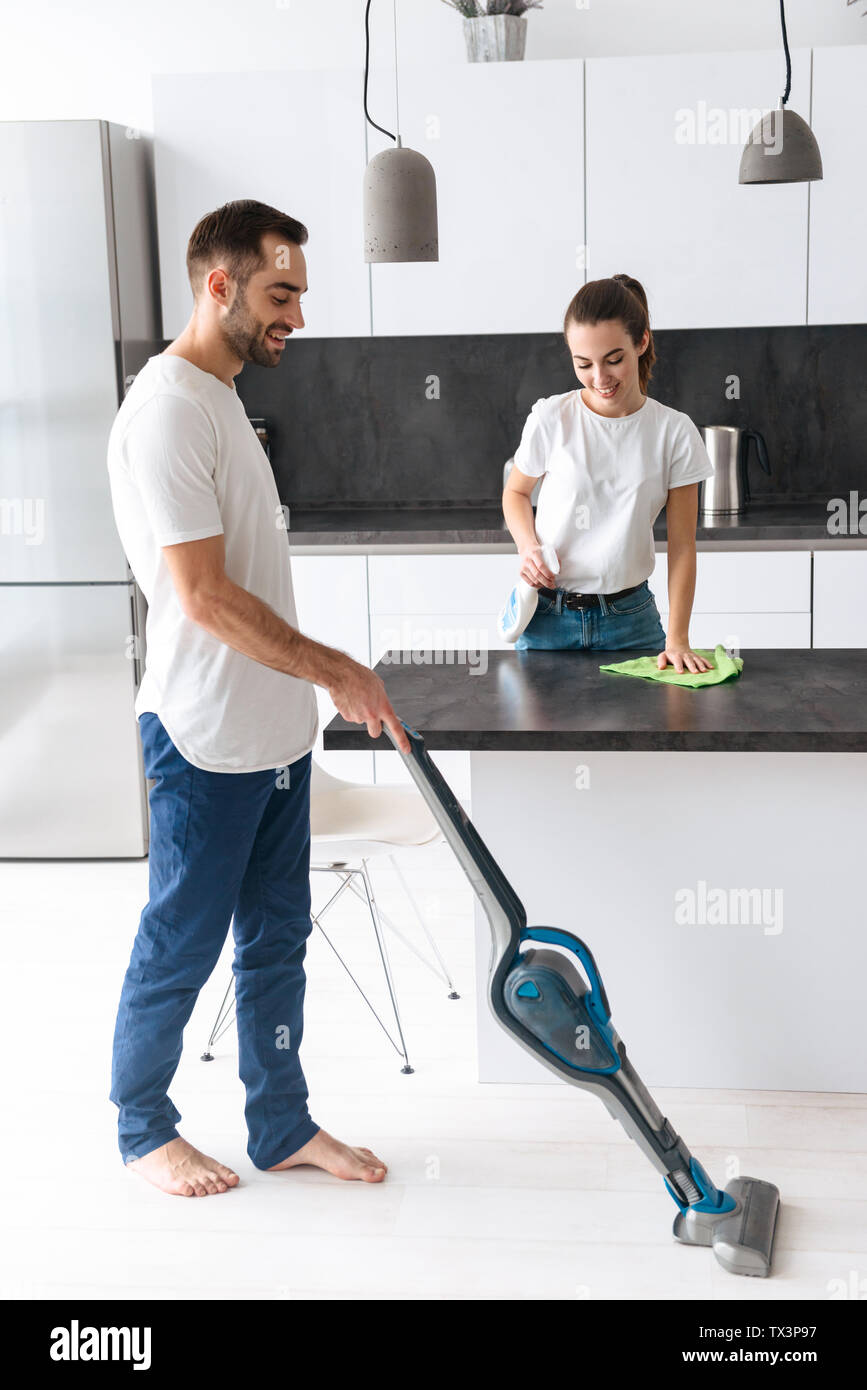 Happy young couple doing general clean at the kitchen, man vacuuming ...