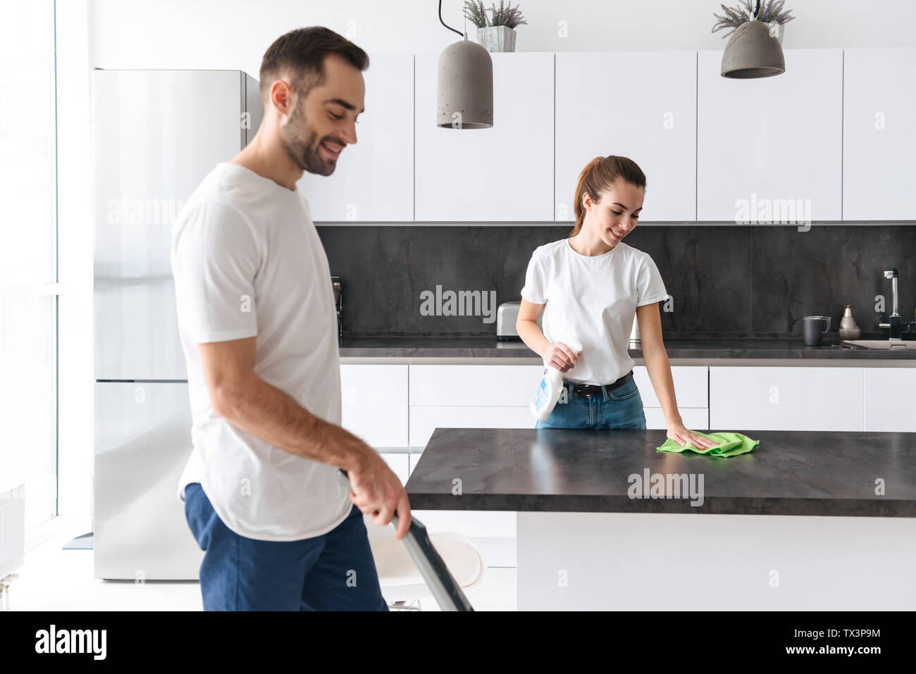 Happy young couple doing general clean at the kitchen, man vacuuming ...