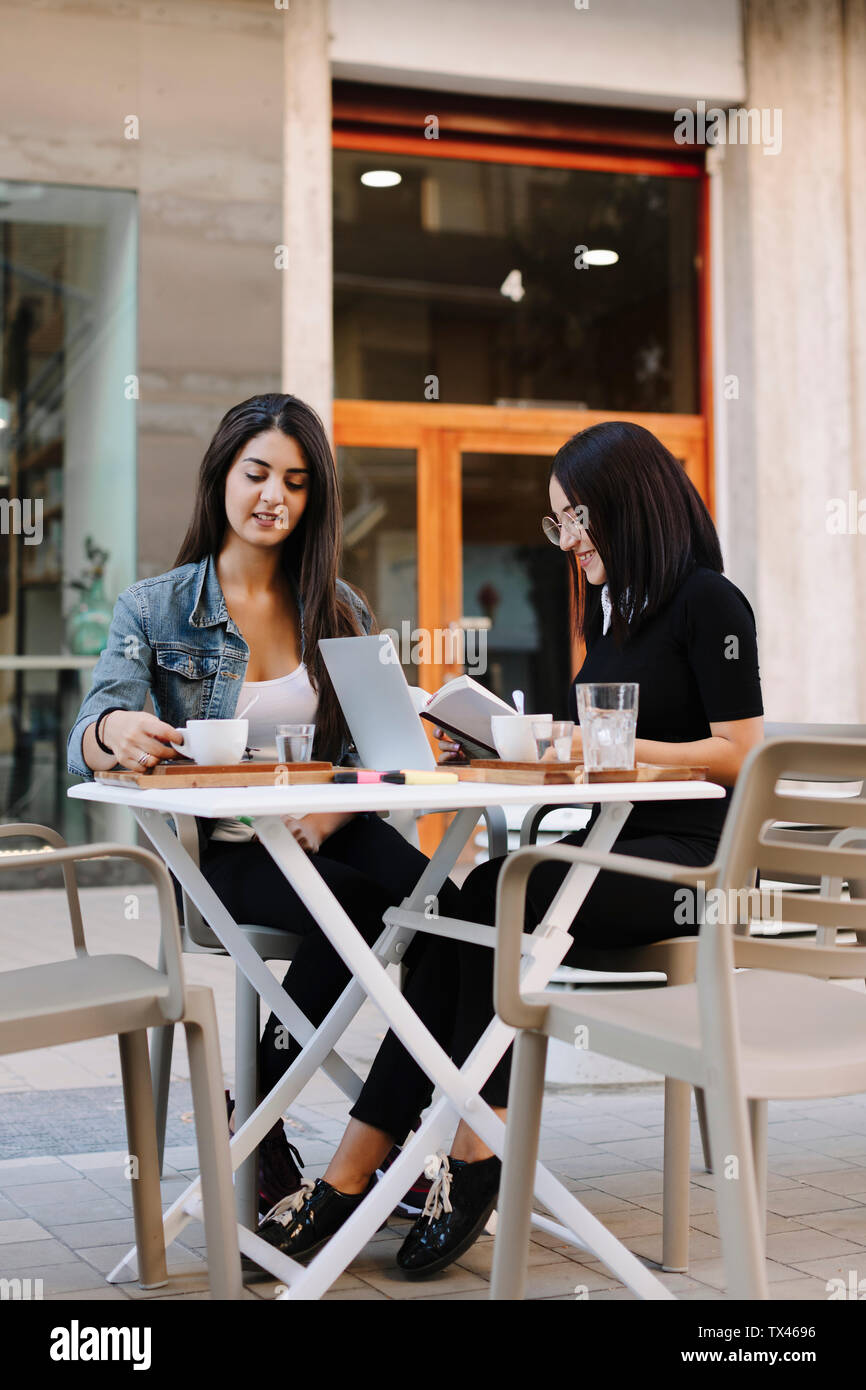 Two friends sitting together at a pavement cafe with book and laptop ...