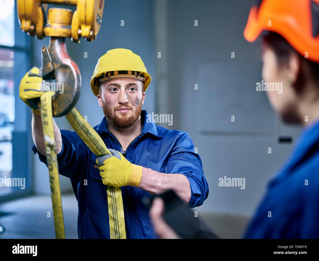 Industrial worker fixing hoist sling on indoor crane, female colleague ...