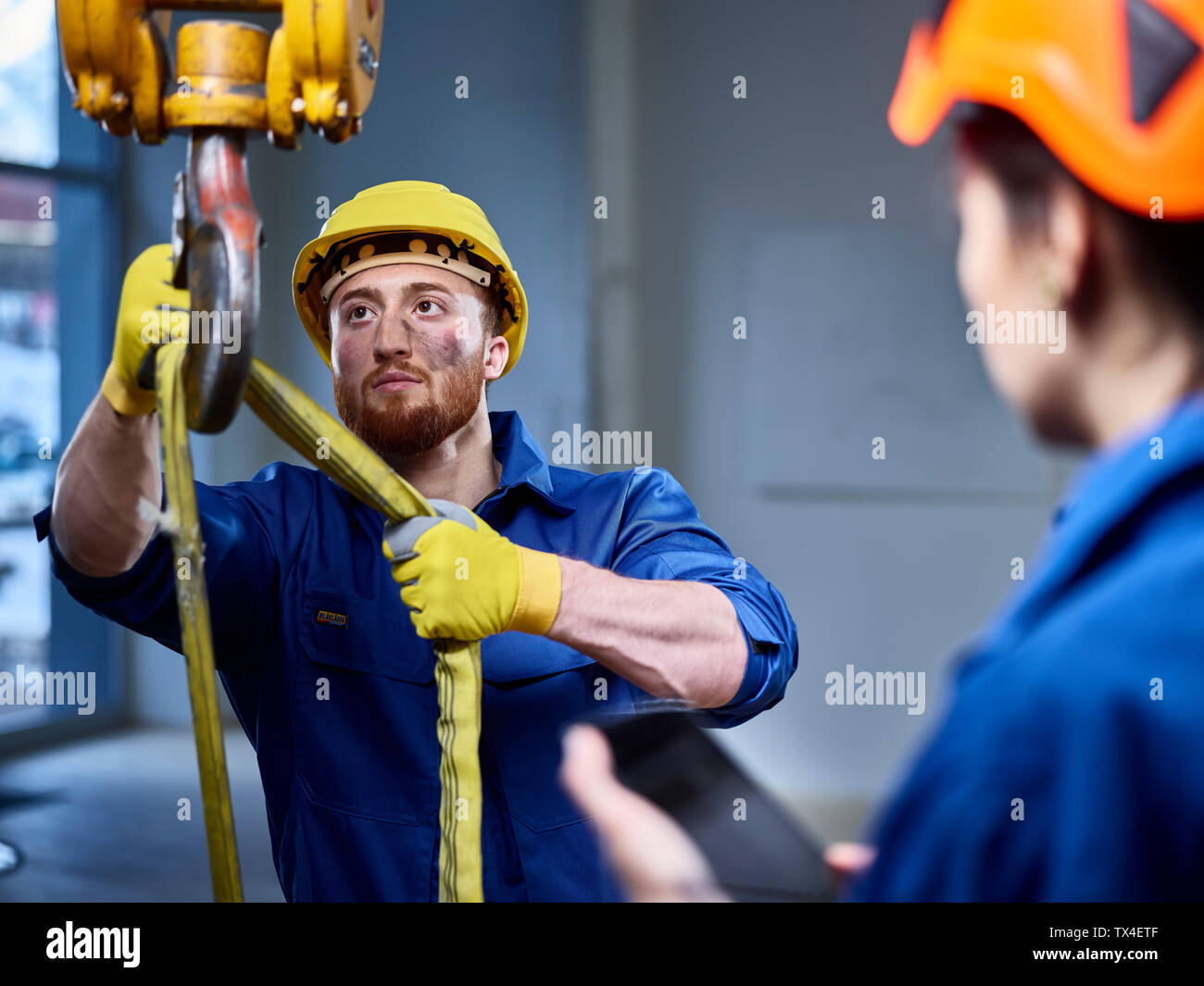 Industrial worker fixing hoist sling on indoor crane, female colleague ...