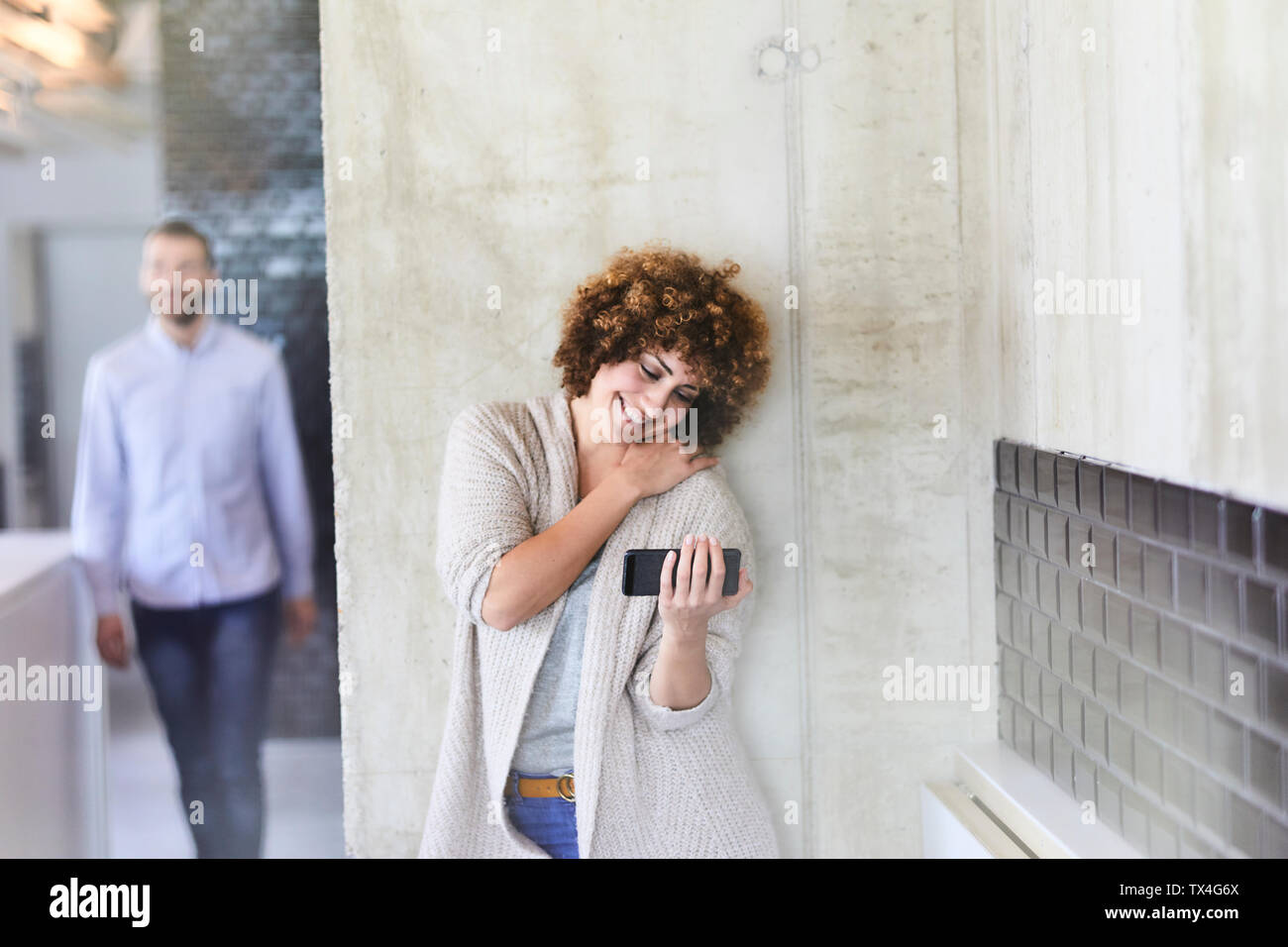 Smiling woman using cell phone at concrete wall with man in background ...