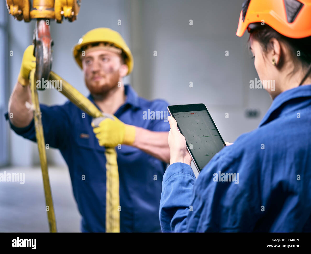 Industrial worker fixing hoist sling on indoor crane, female colleague ...
