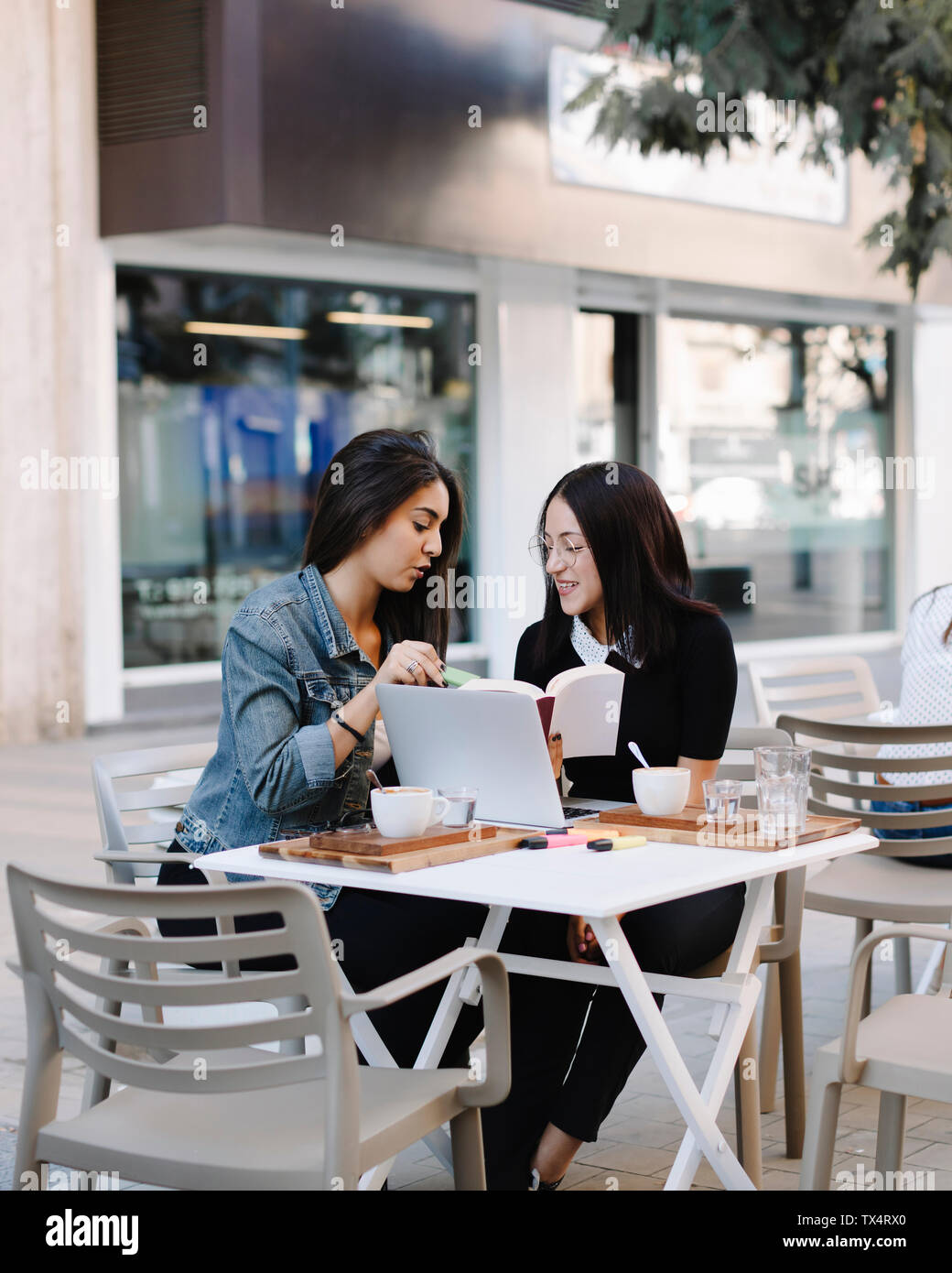 Two friends sitting together at a pavement cafe with book and laptop ...