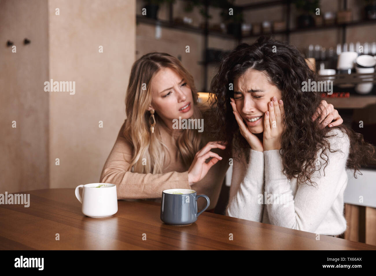 Two upset young girls friends sitting at the cafe table, drinking ...