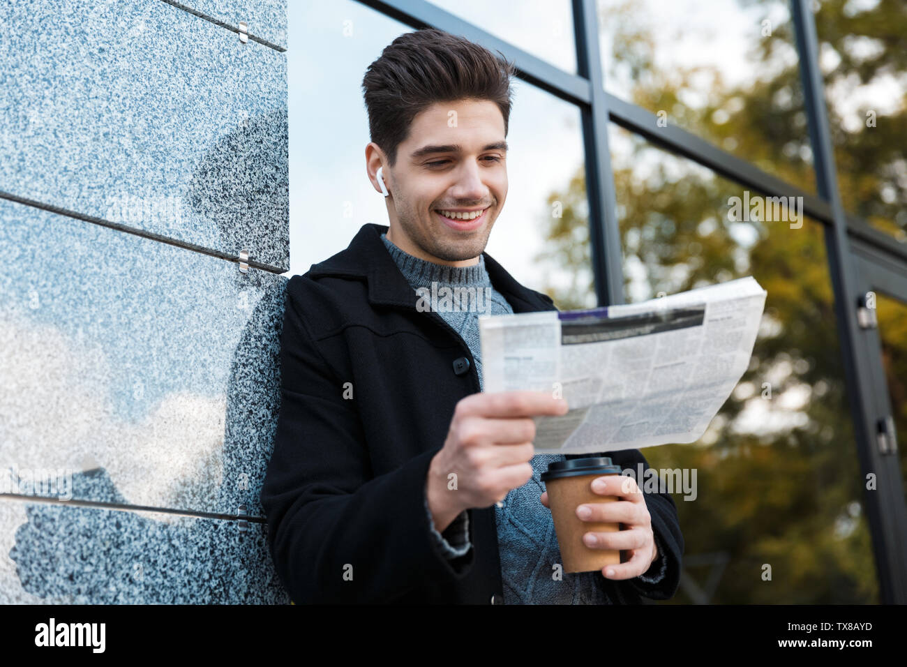 Portrait of cheerful man 30s wearing earpods reading newspaper and ...