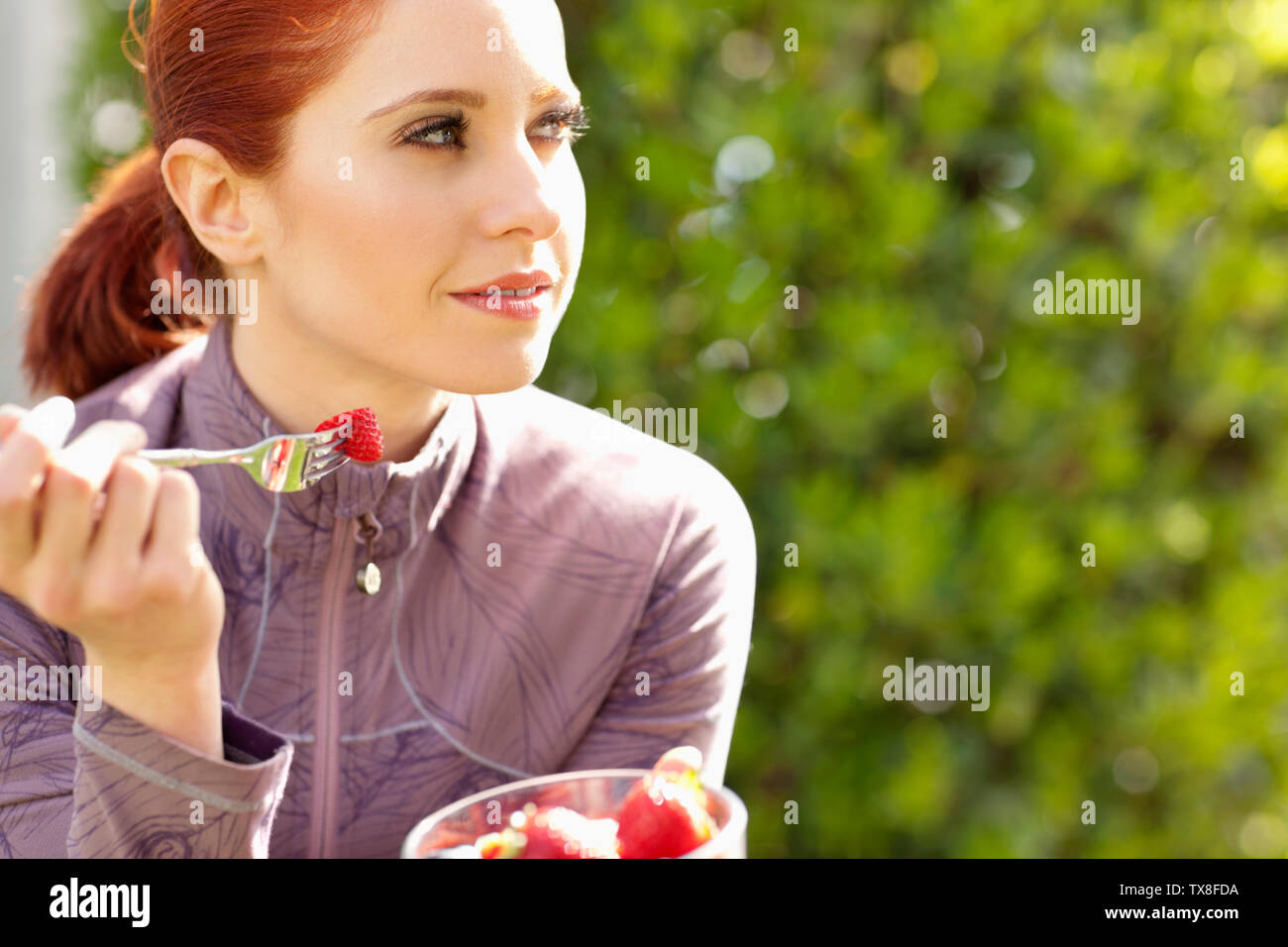 Pretty young woman sits in the sun eating a bowl of strawberries with a ...