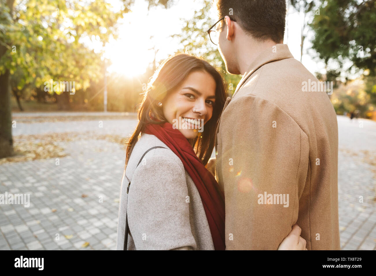 Image of brunette beautiful woman hugging her boyfriend and smiling while walking in autumn park ...